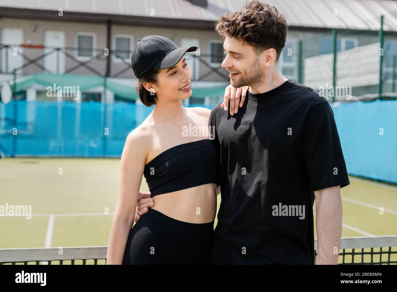 positive couple in black active wear standing near tennis net, romance ...