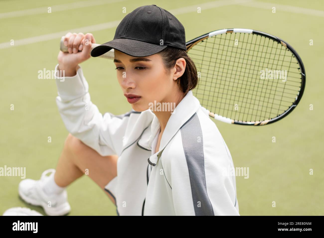 portrait, woman in sportswear and cap sitting on tennis court, holding ...