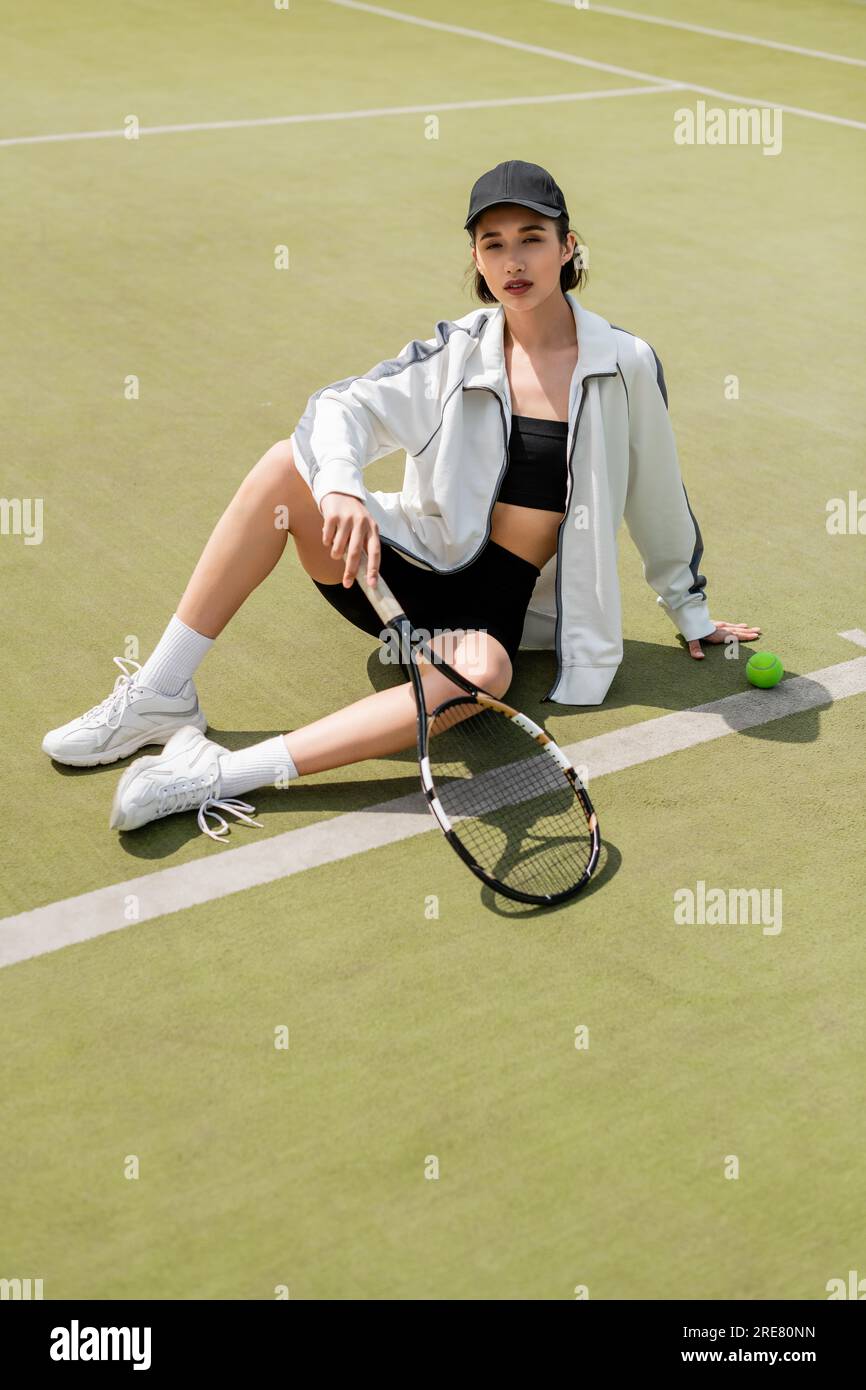 woman in active wear and cap holding tennis racket and sitting on court ...