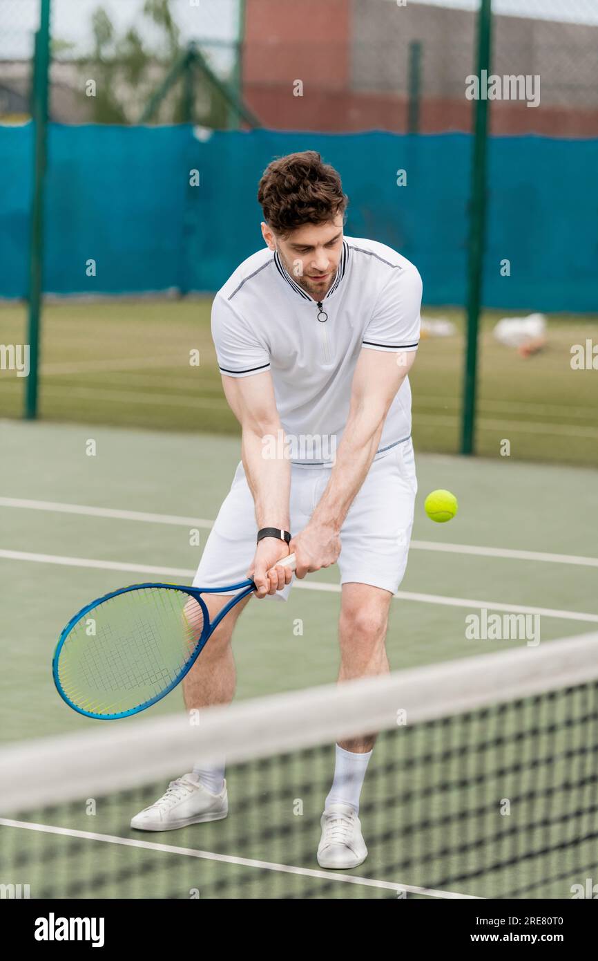 handsome tennis player holding racket and hitting tennis ball on court ...