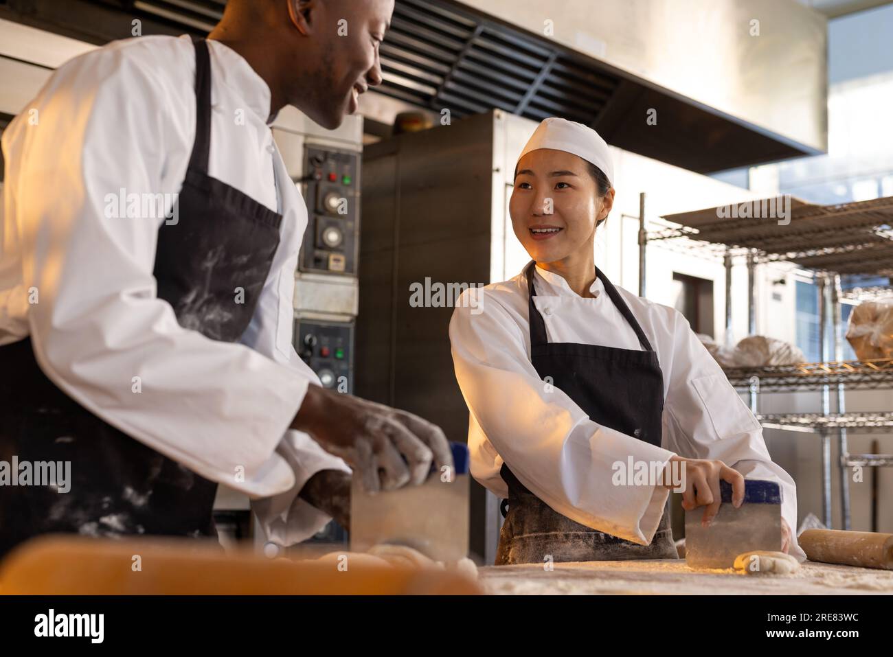 Happy diverse bakers wearing aprons in bakery kitchen and cutting dough ...