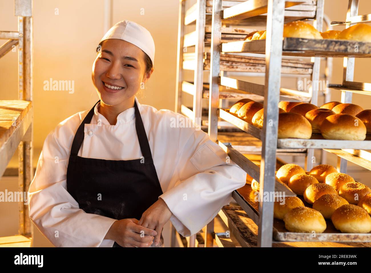Portrait of happy asian female baker in bakery kitchen with fresh rolls ...