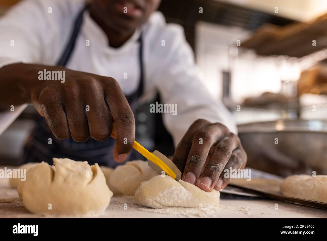 African american male baker in bakery kitchen cutting dough for rolls ...