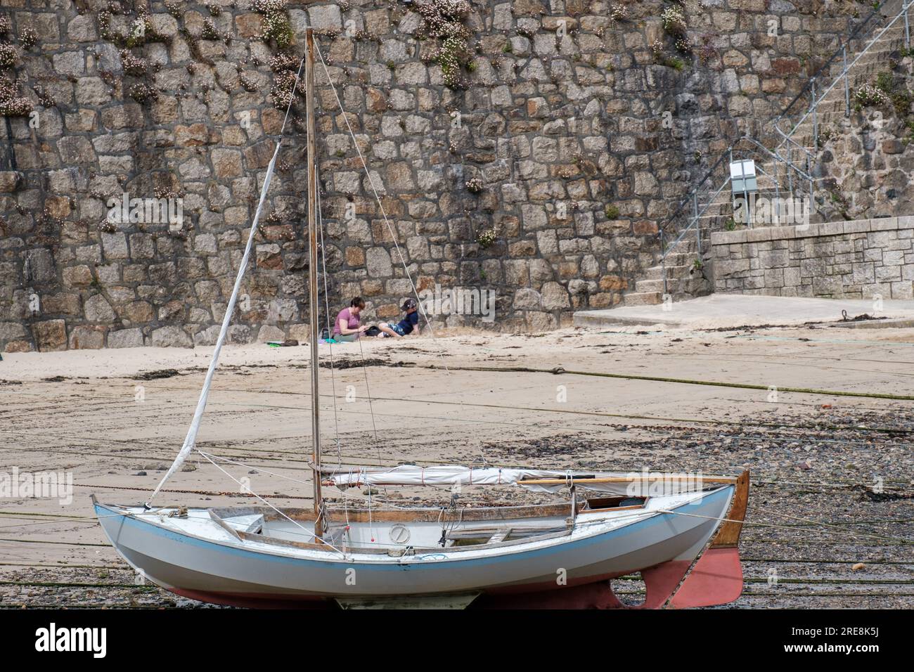 Small sailing boat with sails furled berthed on sand at Mousehole harbour with people sitting on ...