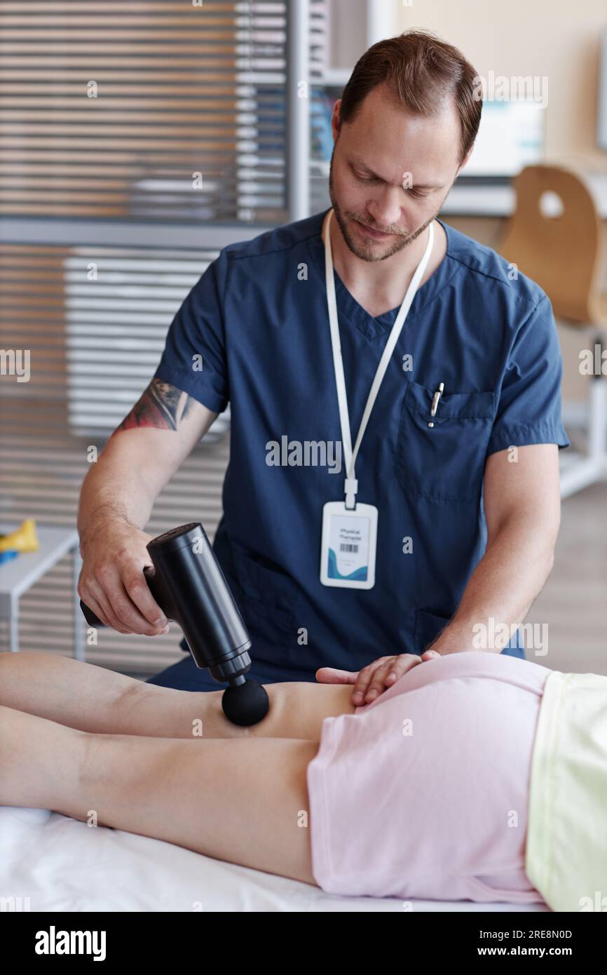 Vertical image of therapist in uniform using massager to massage legs ...