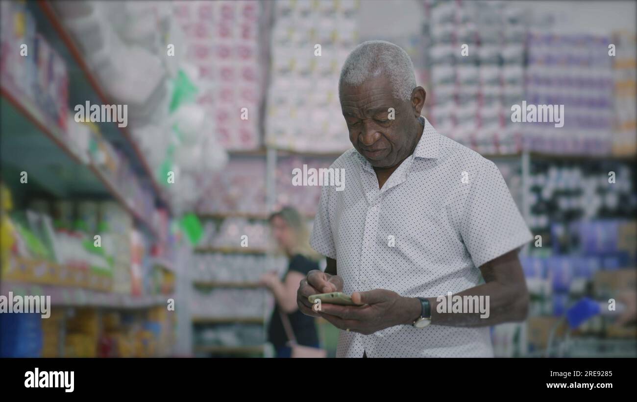 One black senior Brazilian man checking phone standing inside grocery ...