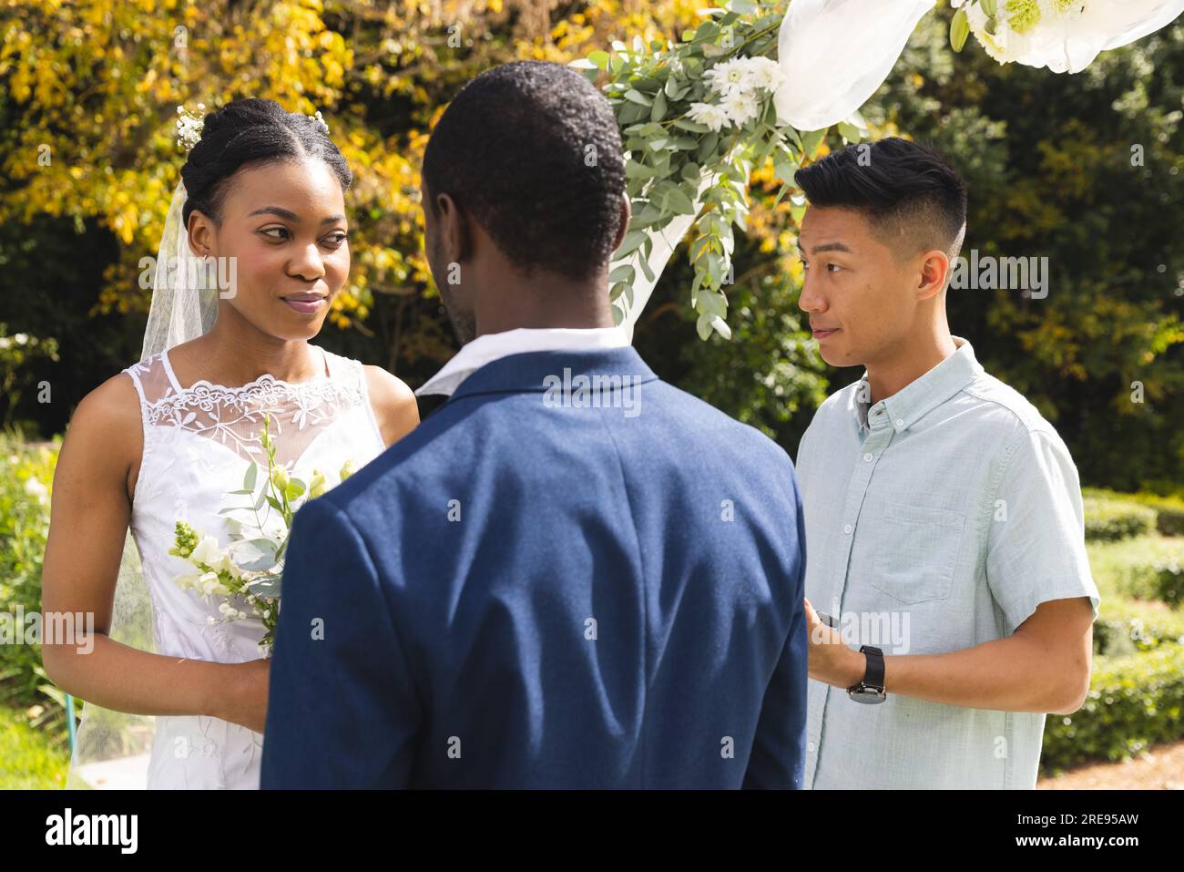 Happy diverse male officiant with bride and groom at outdoor wedding ...