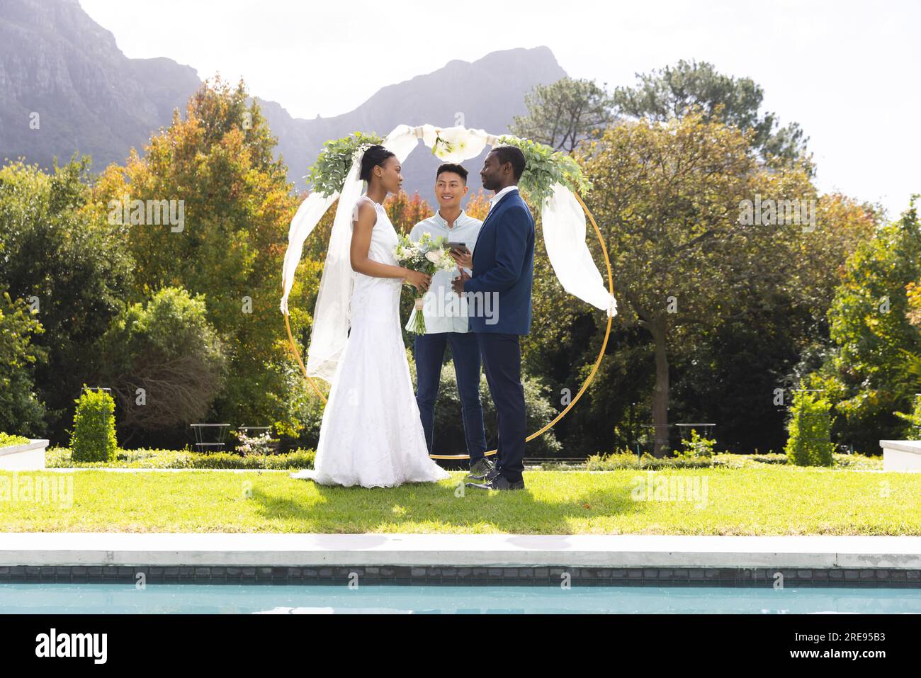 Happy diverse male officiant, bride and groom at outdoor wedding ...
