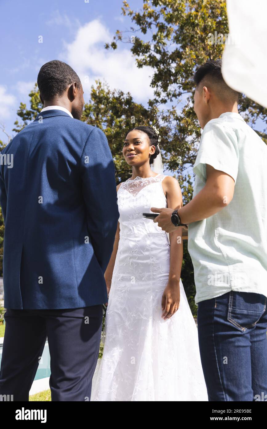 Happy diverse male officiant, bride and groom at outdoor wedding in ...