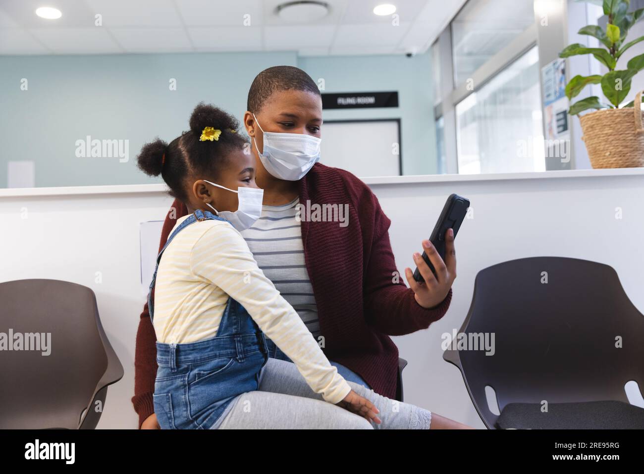 African american mother and daughter wearing face masks using smartphone in waiting room at ...