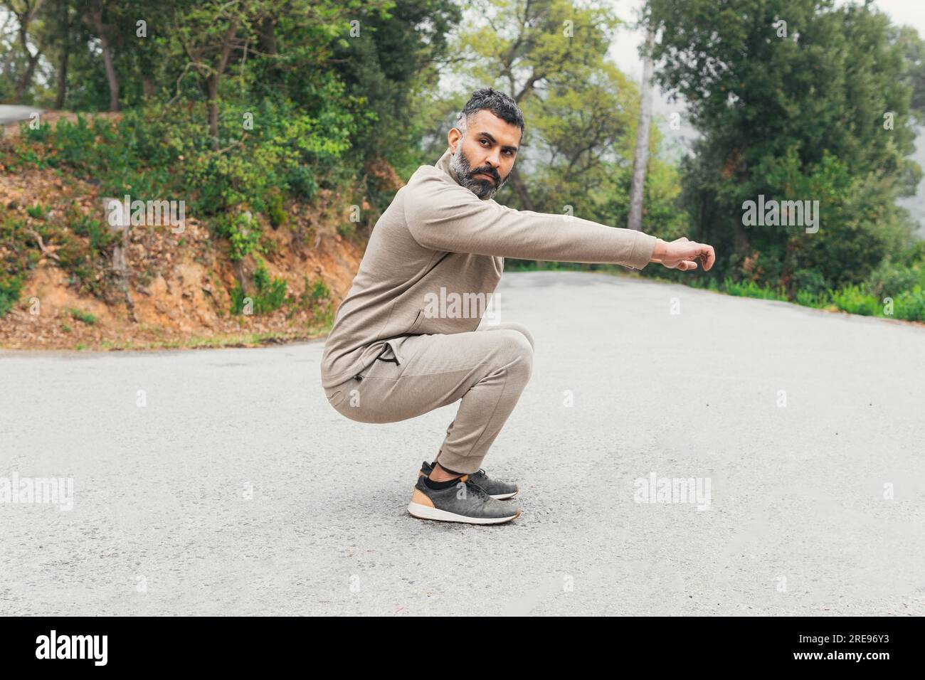 Side view of bearded Indian male looking at camera while squatting on ...