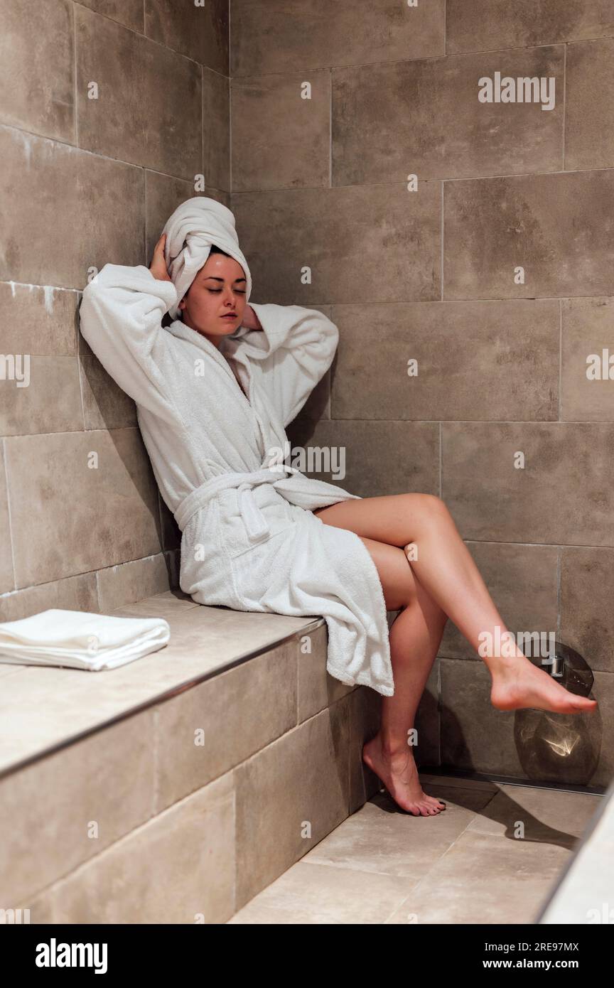 Full body of pensive young female in white bathrobe and touching towel ...