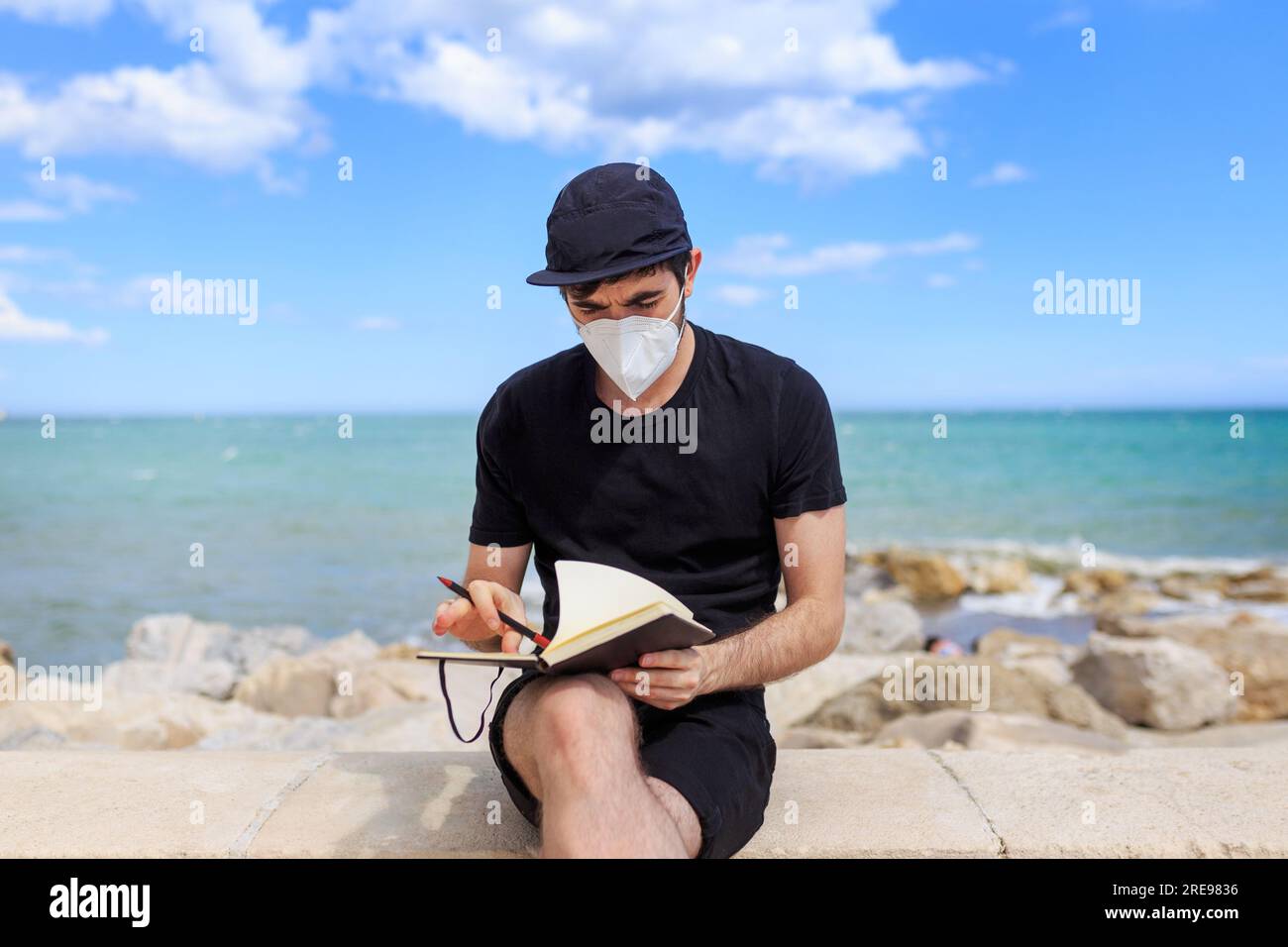 Male author in face mask and cap sitting with crossed legs on fence ...