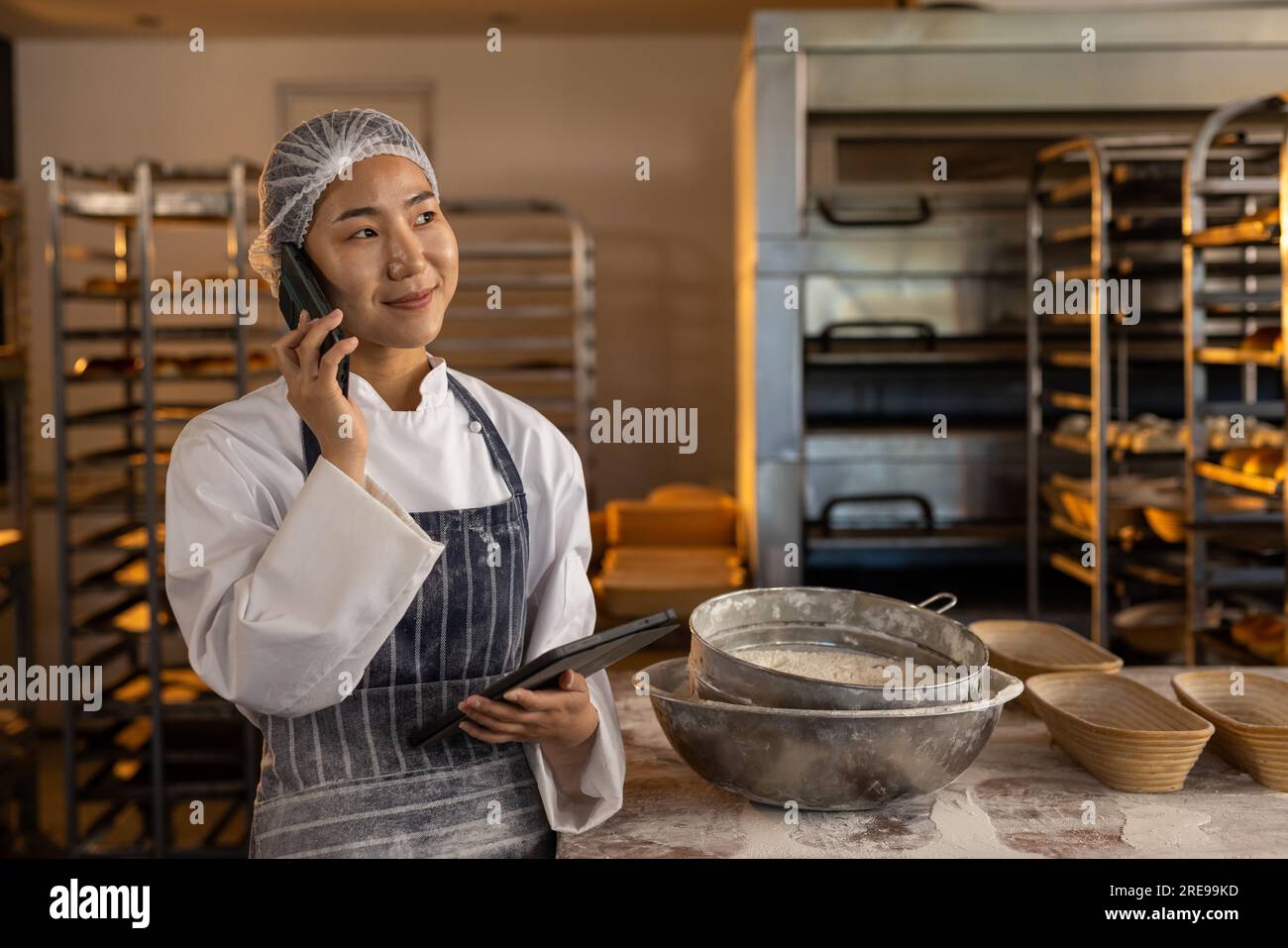 Focused asian female baker in bakery kitchen wearing apron and talking ...