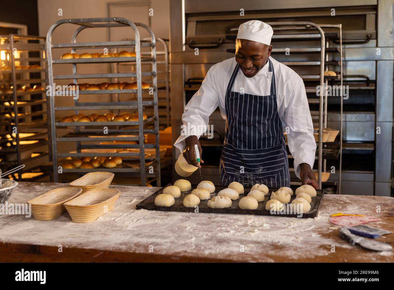 Happy african american male baker in bakery kitchen spraying rolls with ...