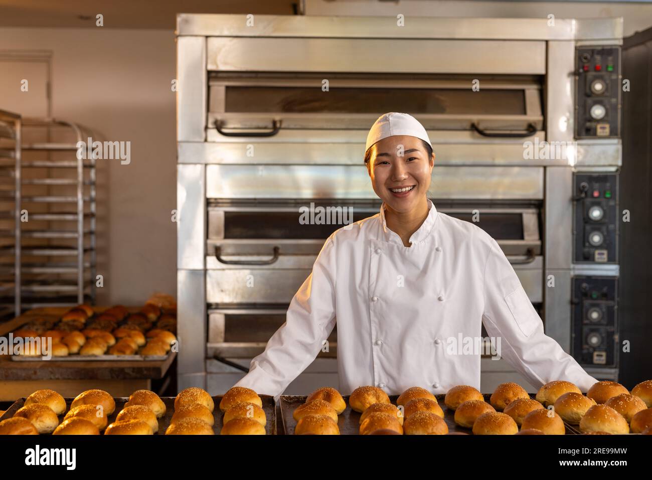 Portrait of happy asian female baker in bakery kitchen with fresh rolls ...
