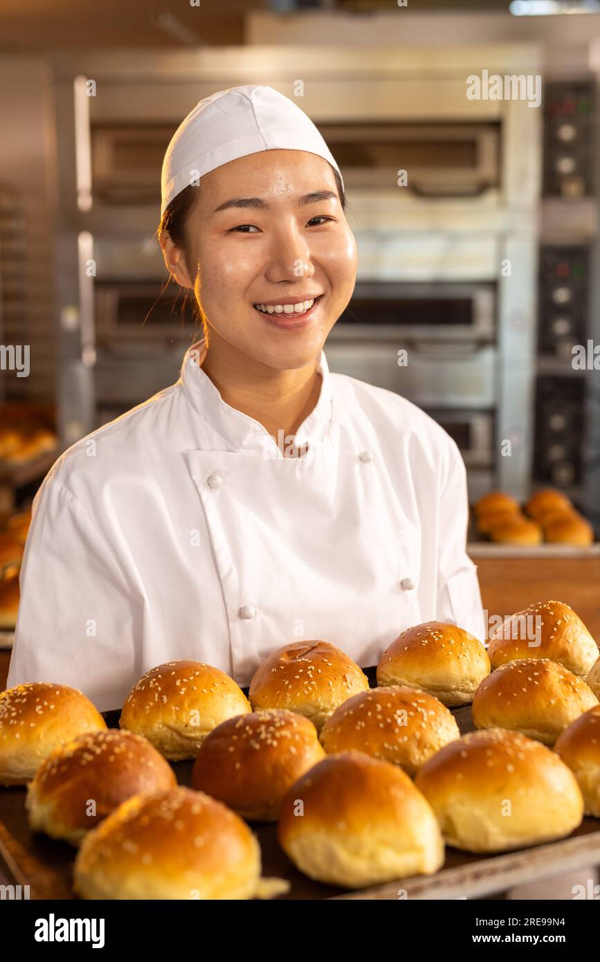 Portrait of happy asian female baker in bakery kitchen presenting fresh ...