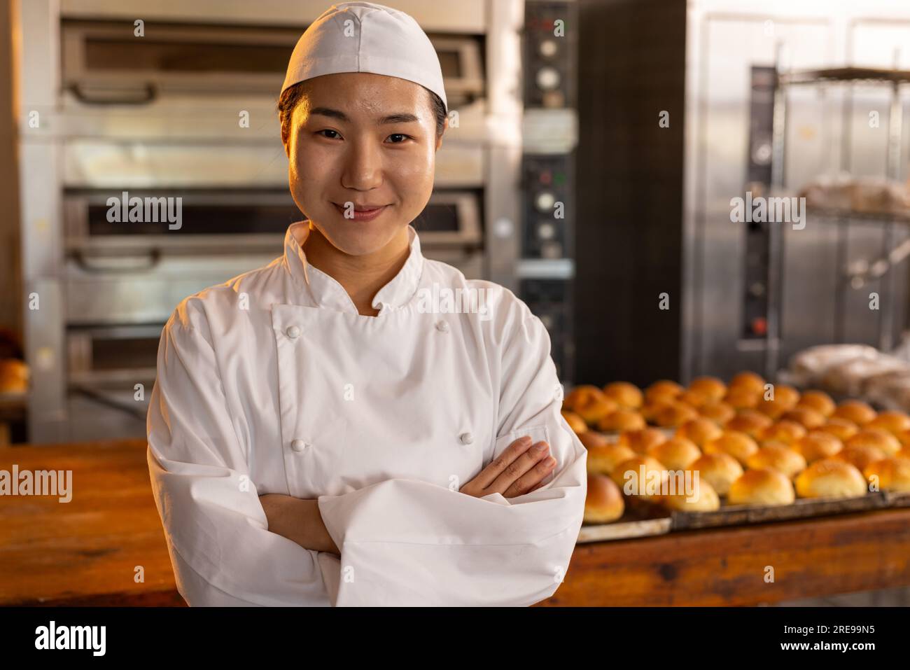 Portrait of happy asian female baker with arms crossed in bakery ...