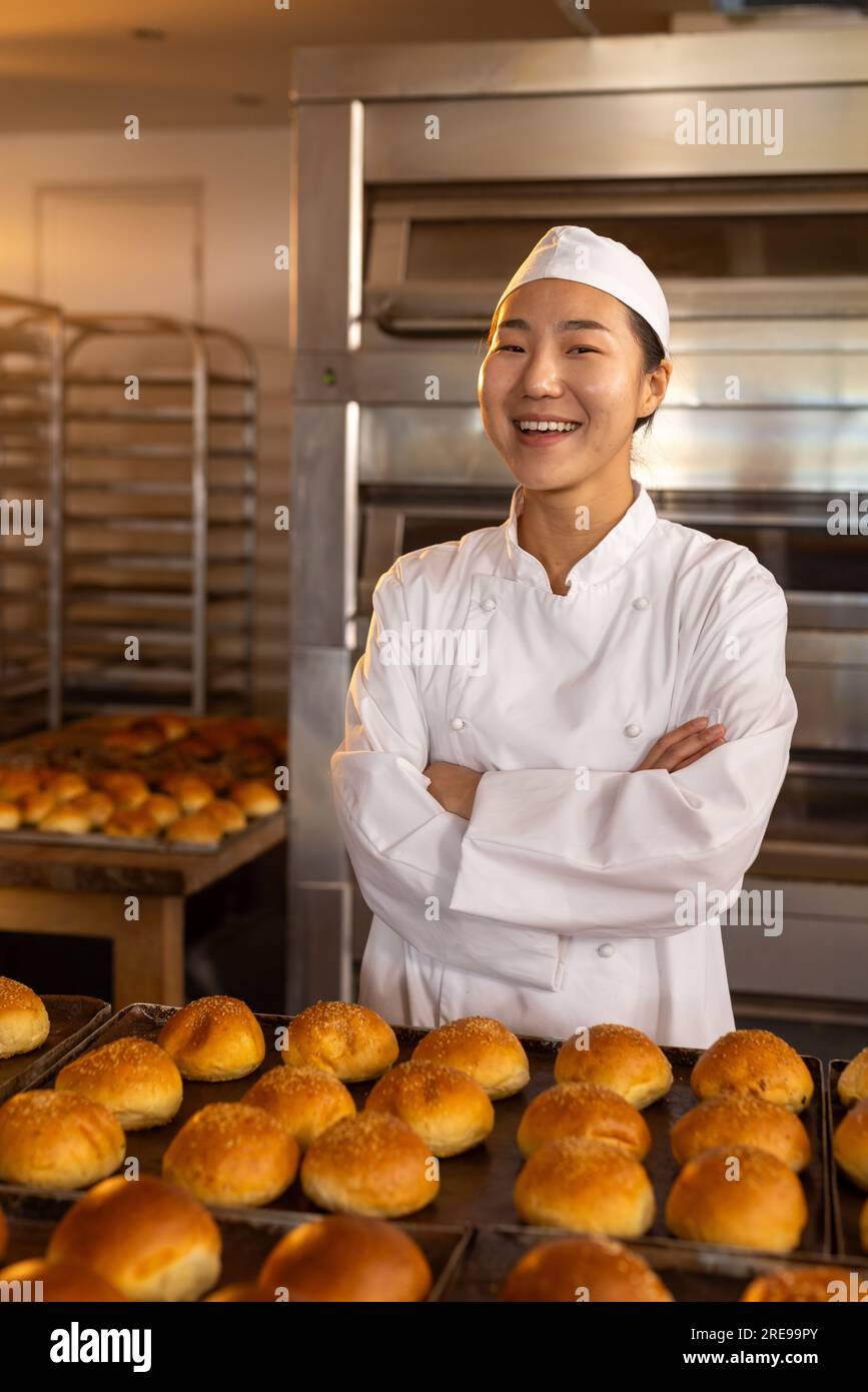 Portrait of happy asian female baker in bakery kitchen with arms ...