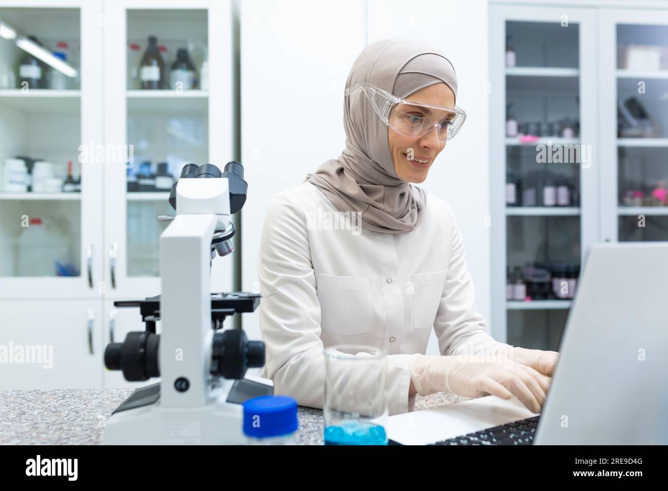 Arab young woman scientist, pharmacist in hijab sitting at table with microscope, liquids in ...