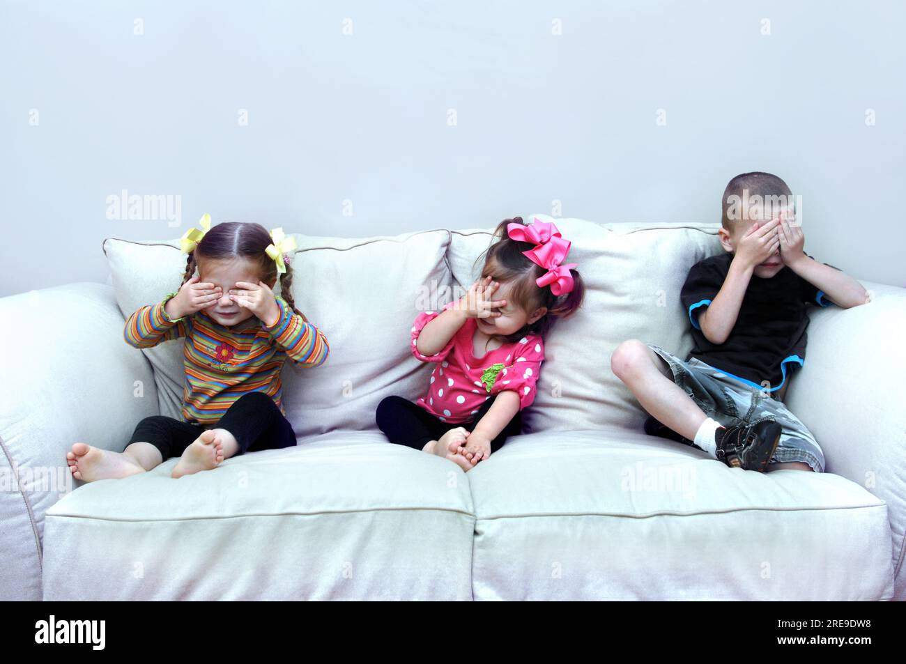 Three small children, sit on a couch in their home, and pretend to see ...
