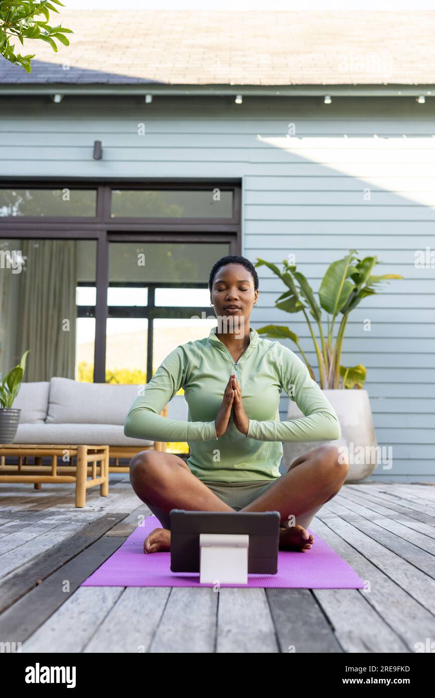 African american woman doing yoga, meditating using tablet on terrace ...