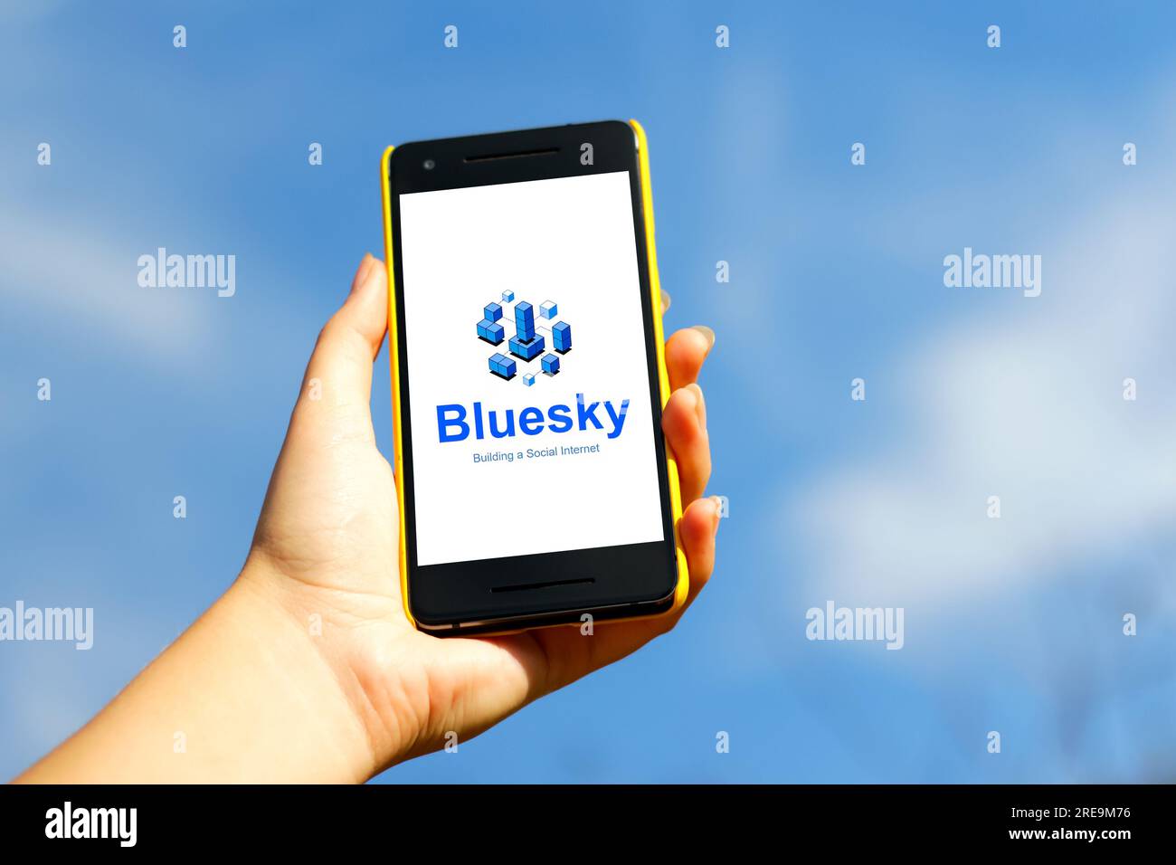 Bluesky app. Hand holding smartphone in yellow plastic case on blue sky ...