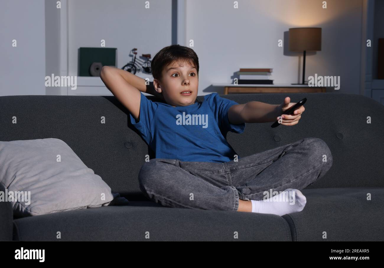 Emotional boy changing TV channels with remote control on sofa at home ...