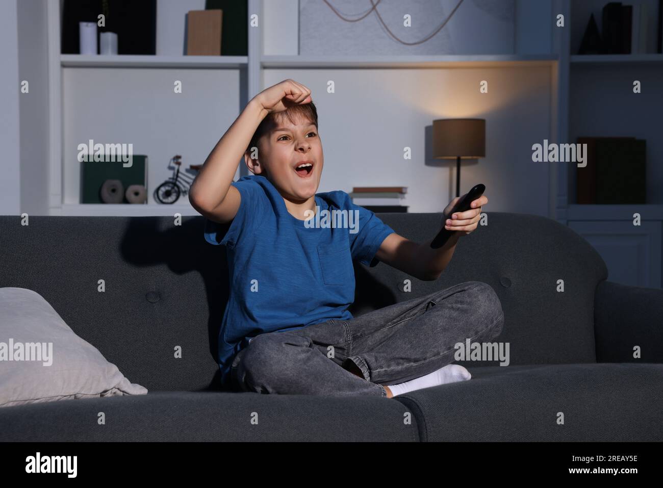 Emotional boy watching TV and holding remote control on sofa at home ...
