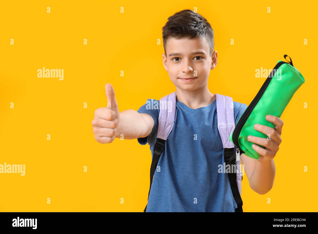 Little schoolboy with pencil case showing thumb-up on orange background ...