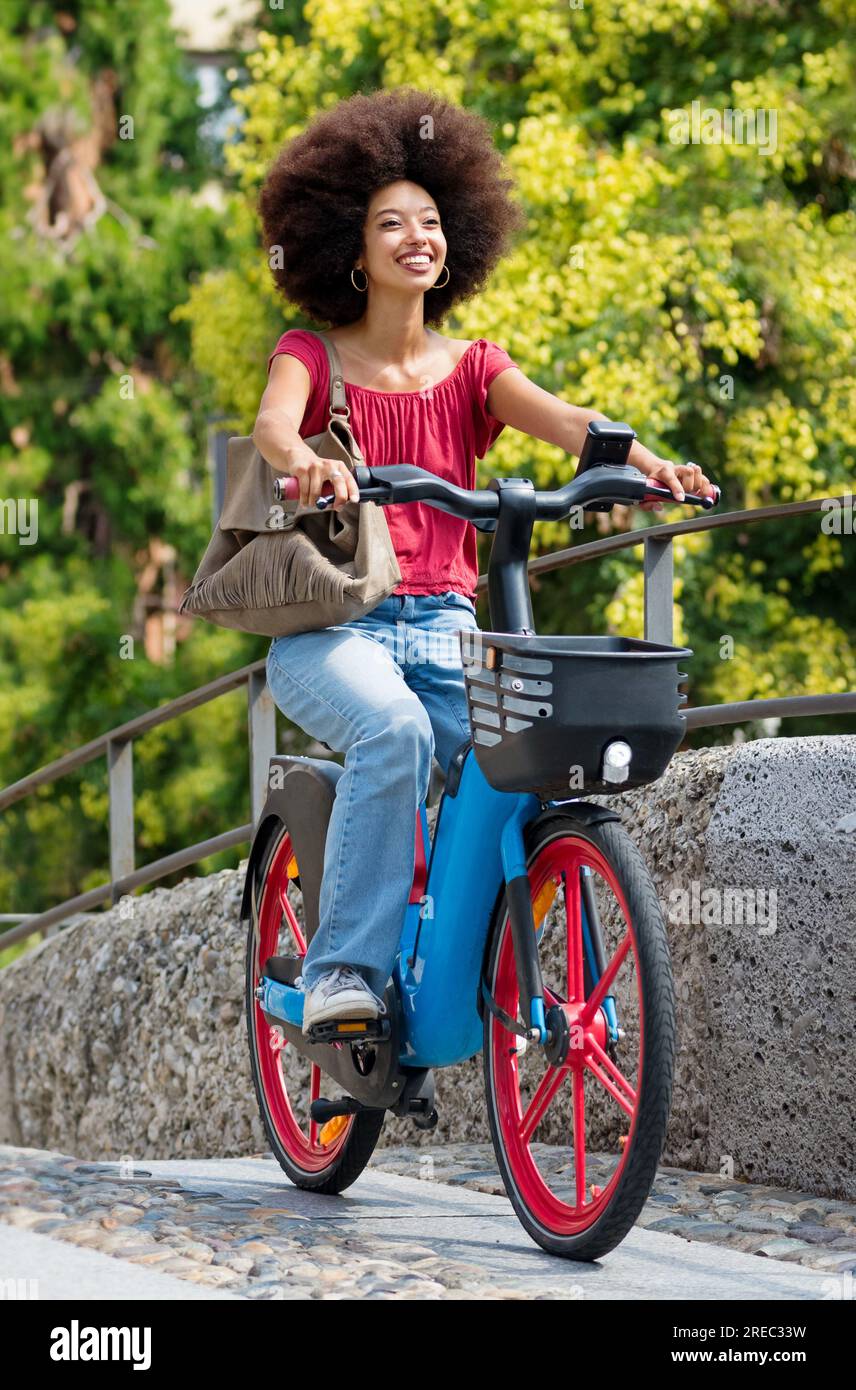 Full length of satisfied black lady with Afro hairstyle riding bicycle and looking away on urban ...
