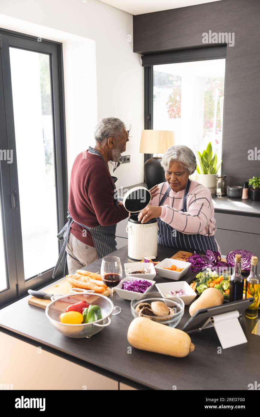 Senior biracial couple wearing aprons composting food waste in kitchen ...