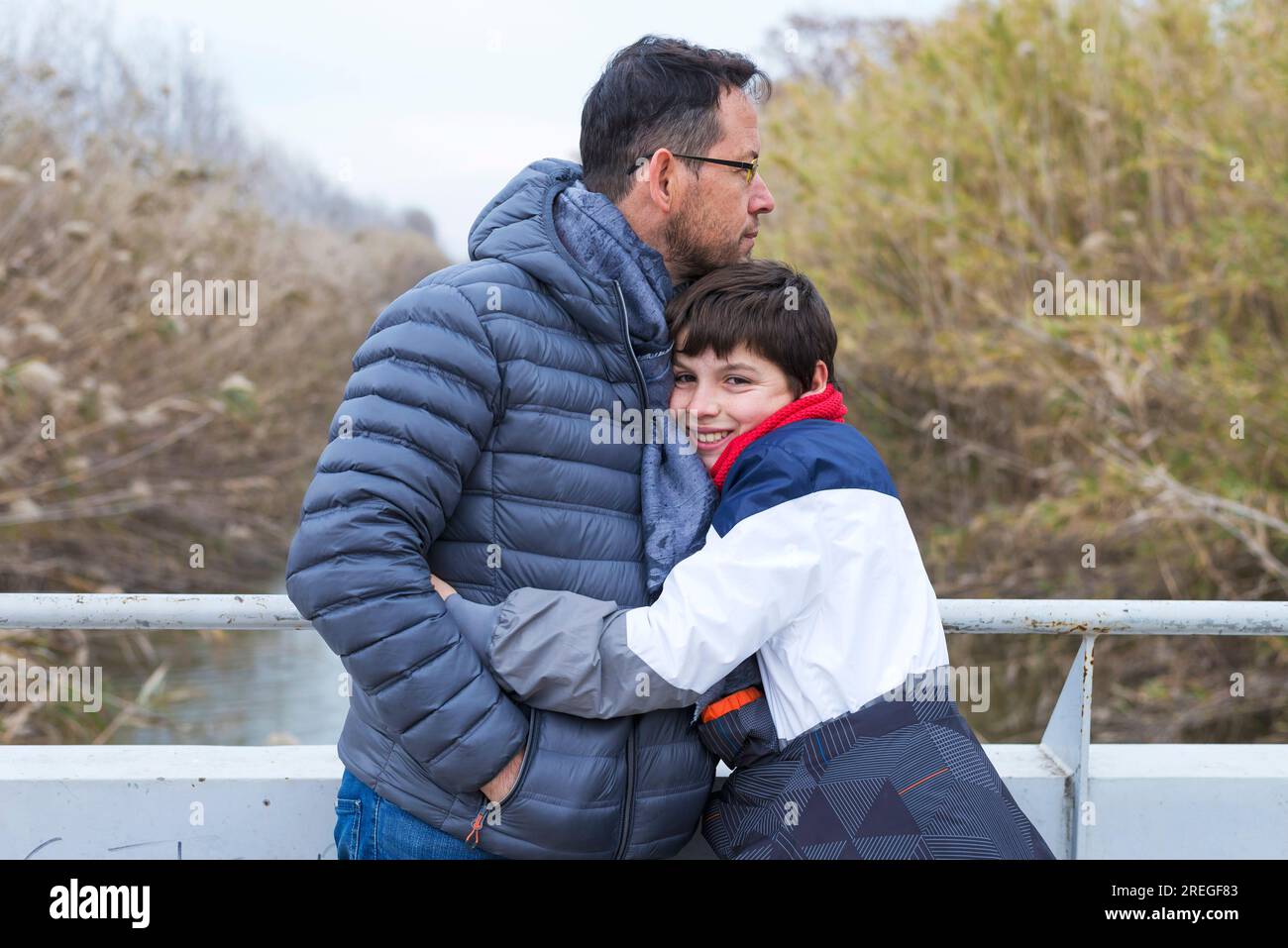 Side view of a father and son hugging while leaning on a railing fence ...