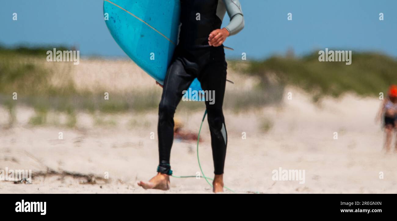 Man walking in a wetsuit carrying his surfboad on the beach with sand ...