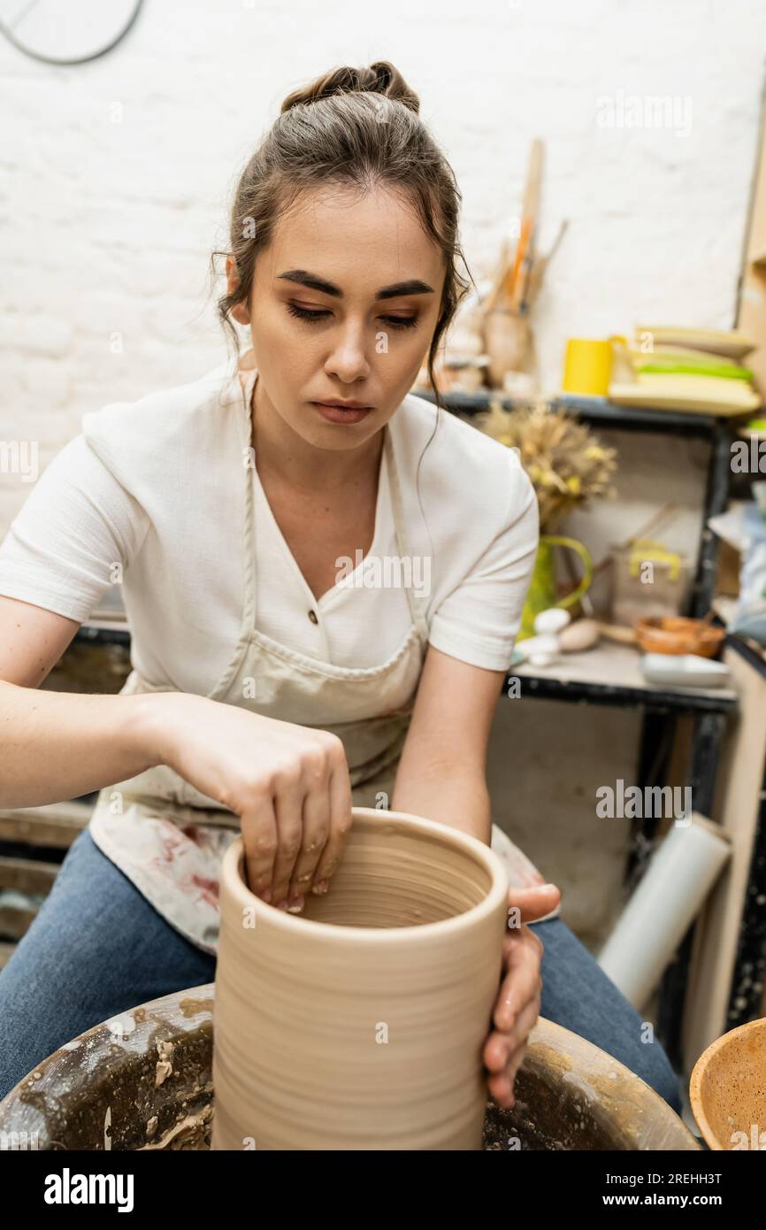 Brunette craftswoman in apron shaping clay on pottery wheel in ceramic ...