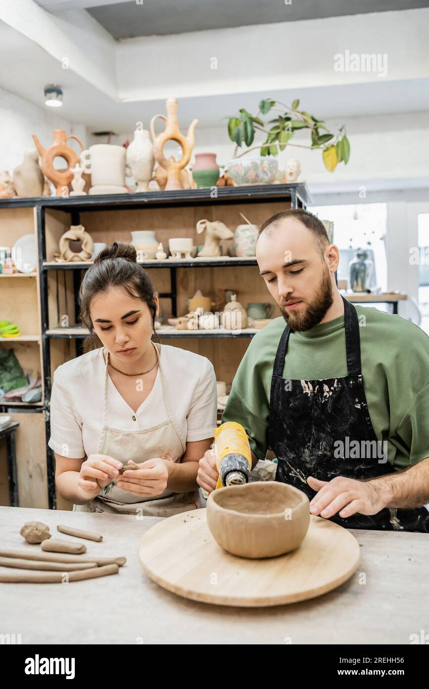 Craftsman drying earthenware with heat gun while girlfriend shaping ...