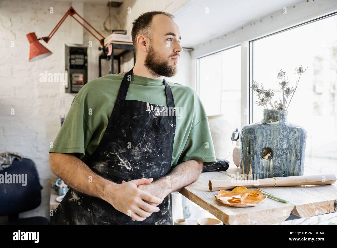 Bearded craftsman in apron looking away while standing near vase with ...