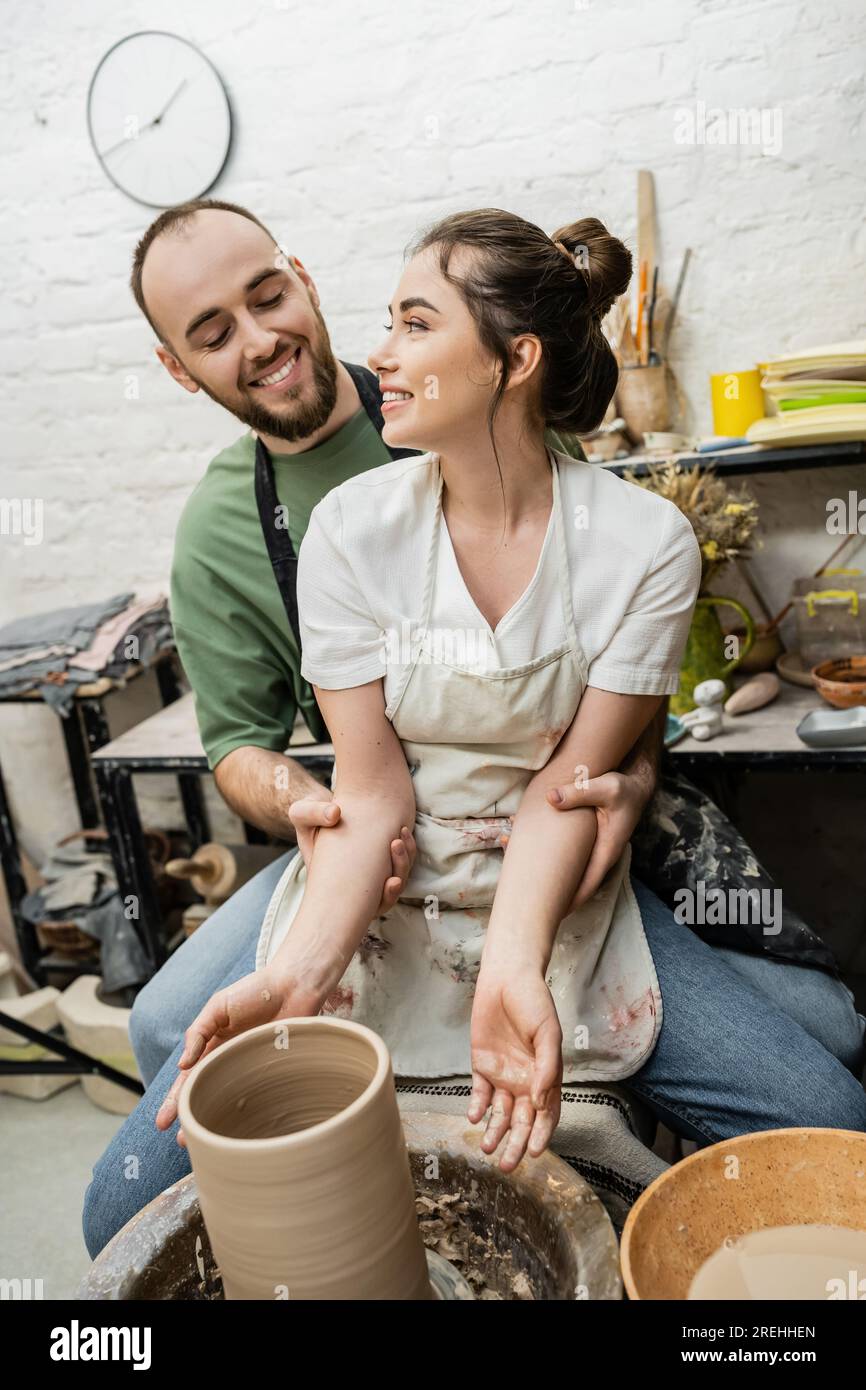 Positive artisan hugging girlfriend in apron and making clay vase on ...