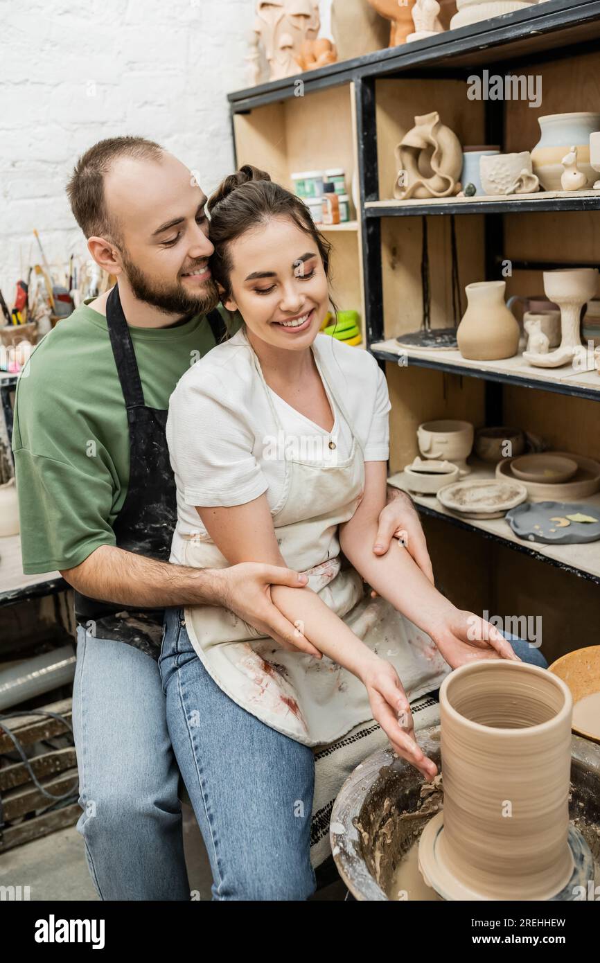 Smiling artisan hugging girlfriend while making clay vase on pottery ...