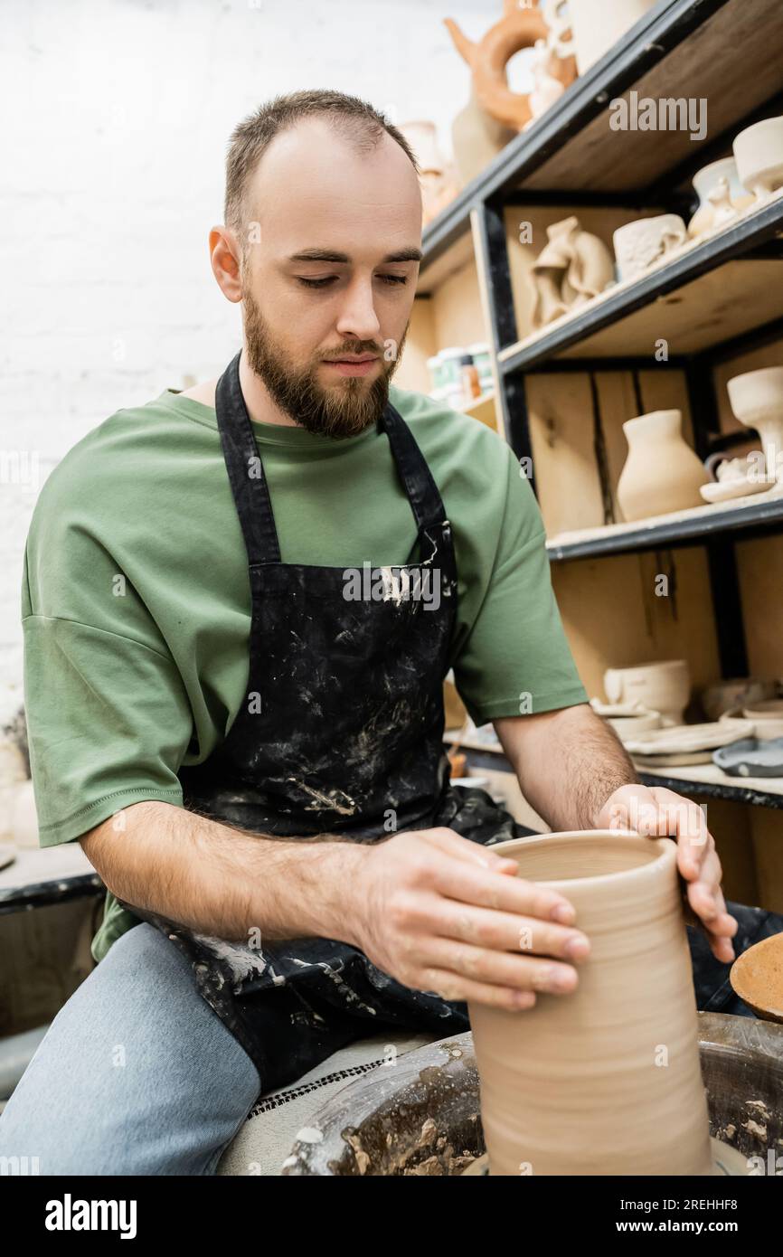 Bearded craftsman in apron shaping clay vase on pottery wheel in ...