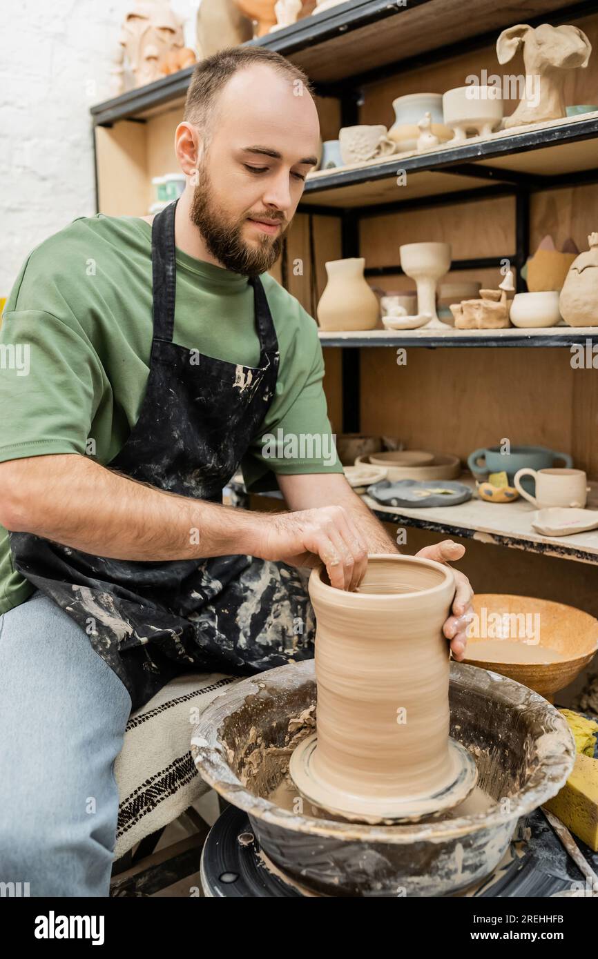Bearded craftsman in apron making clay vase on pottery wheel near rack ...