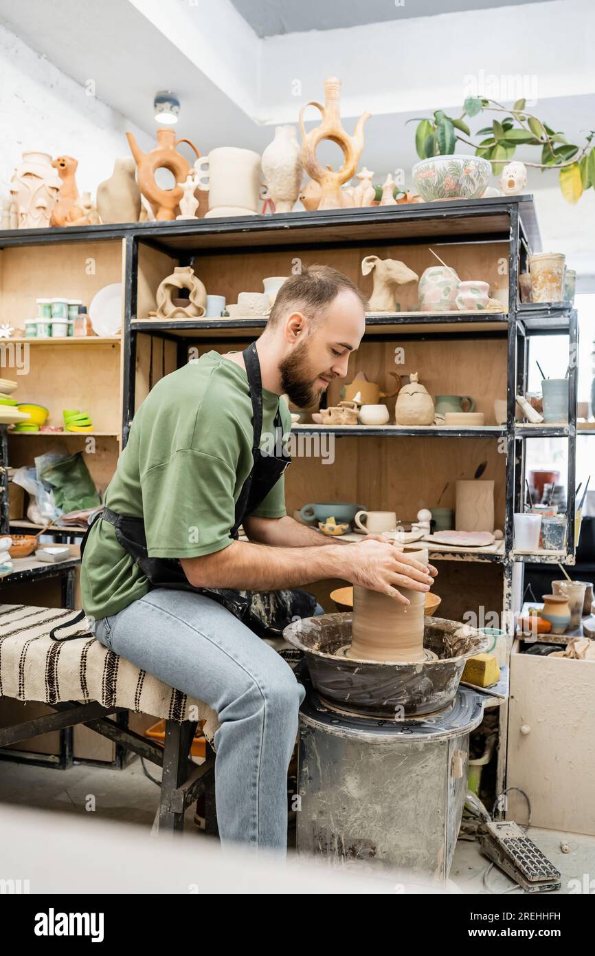 Side view of potter in apron shaping clay sculpture on pottery wheel ...