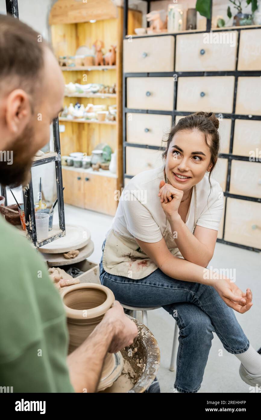 Positive craftsman in apron talking to girlfriend making clay vase on ...