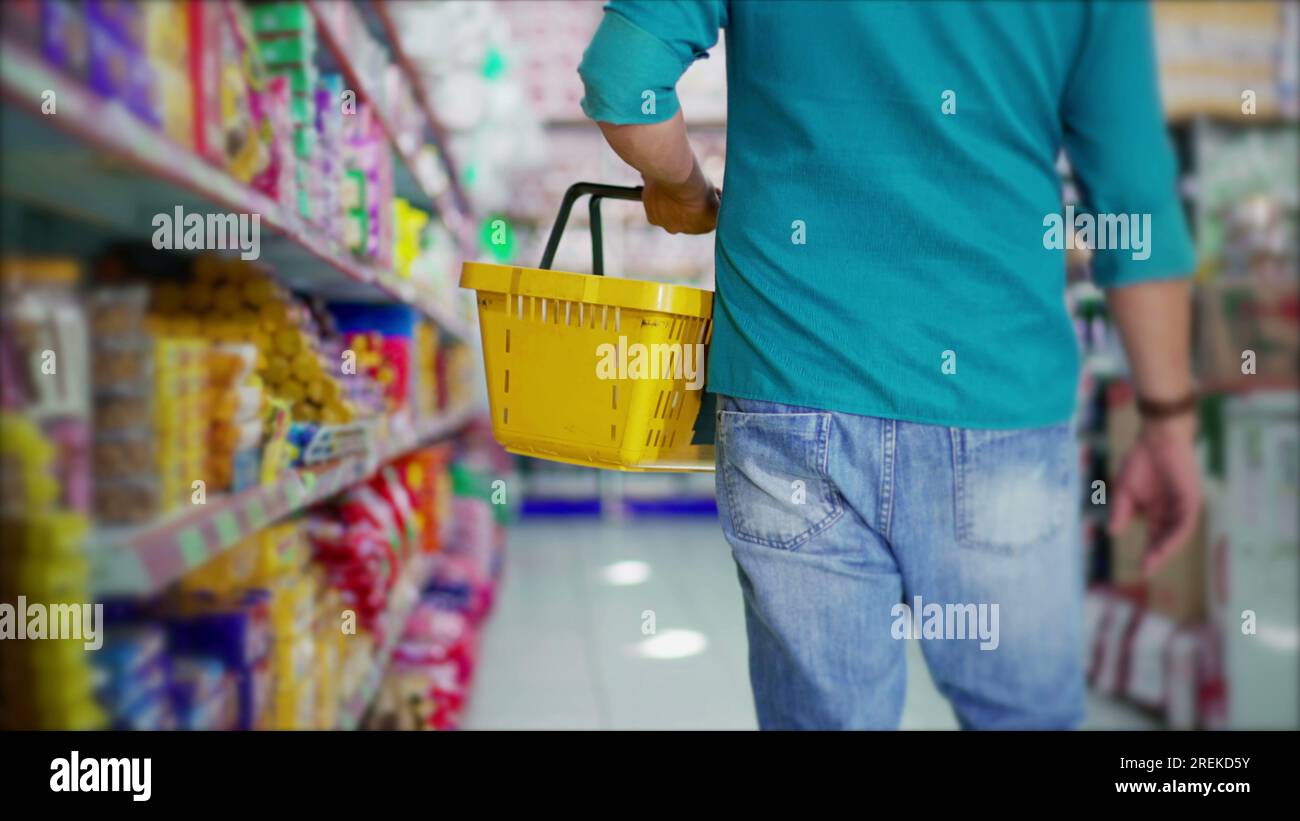 Back of shopper walking in supermarket aisle holding basket in hand ...
