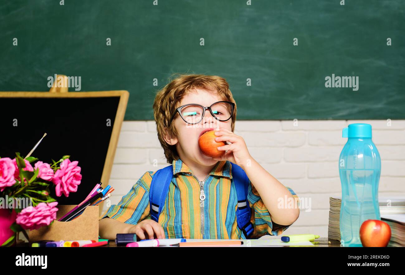 Little schoolboy in glasses sitting at desk eating apple at lunch time ...
