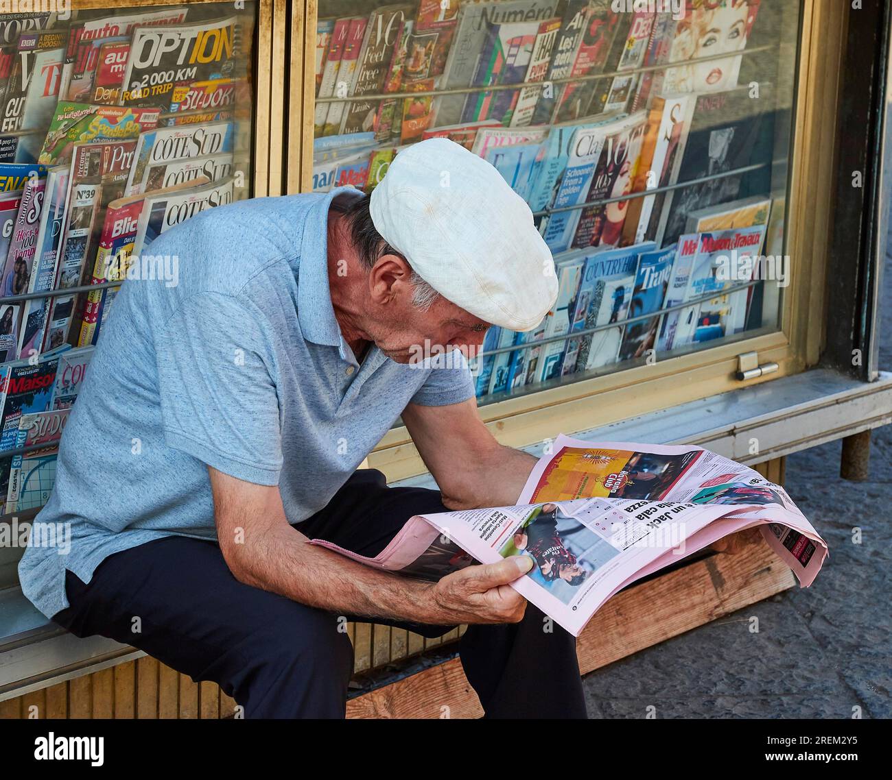 Older man sitting in front of kiosk reading a daily newspaper, white ...
