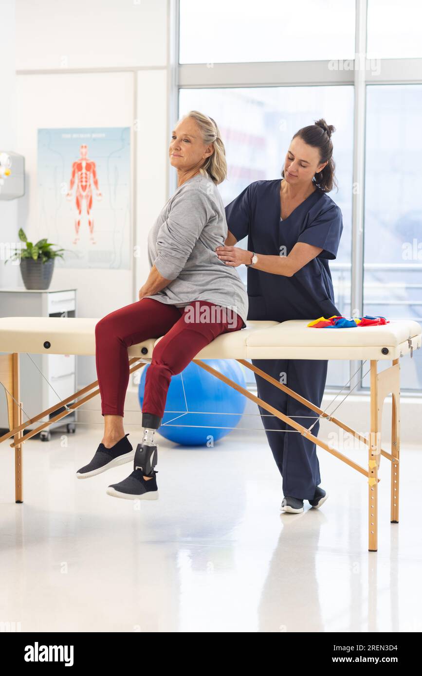 Caucasian female physiotherapist and senior woman with artificial leg massaging back at hospital ...