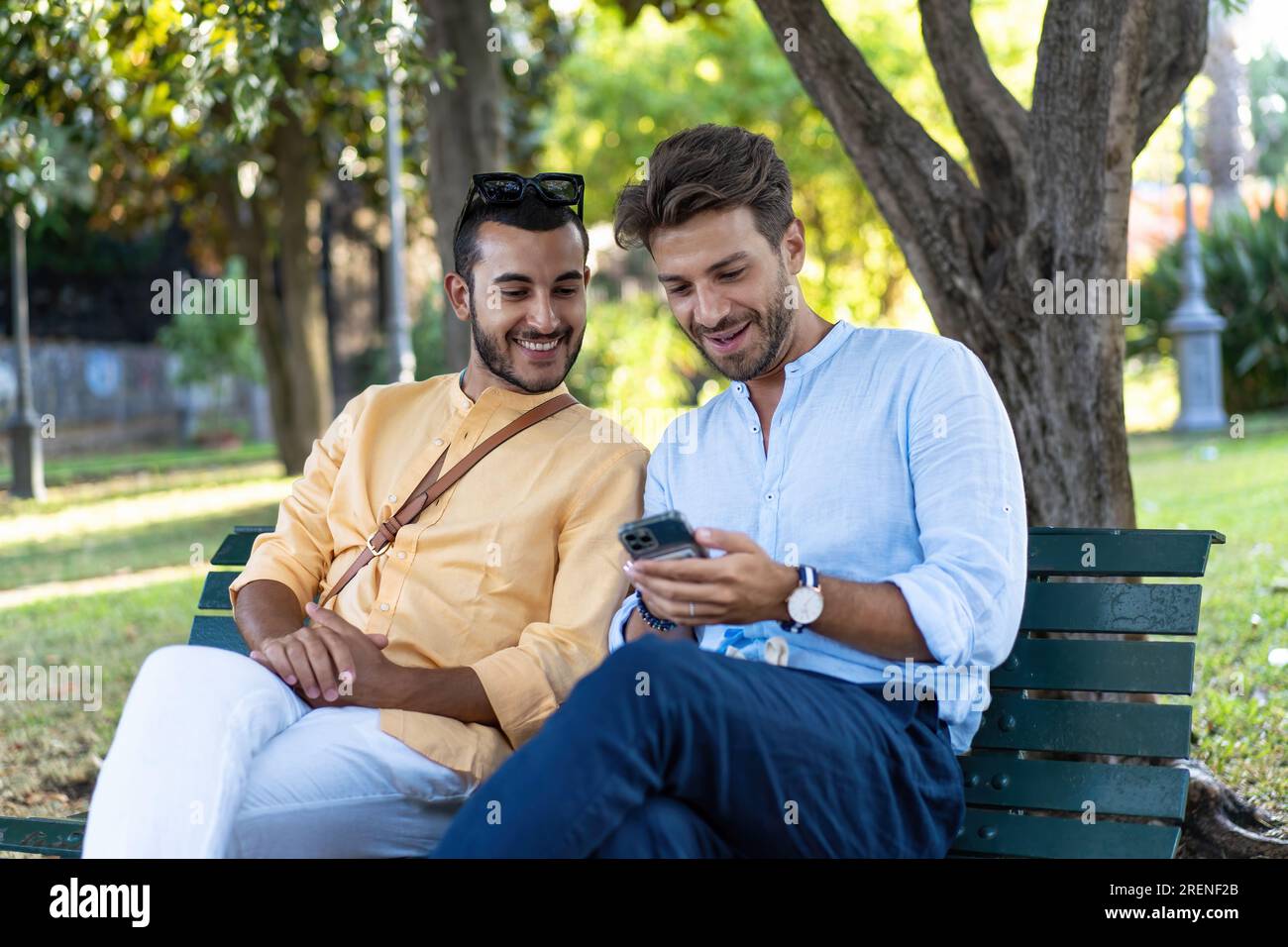 The same two young men sit on a park bench, laughing while viewing ...