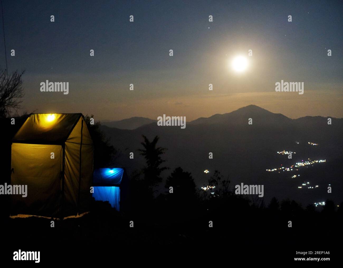 Lit-up camps on a campground with a rising full moon on a hill top in ...