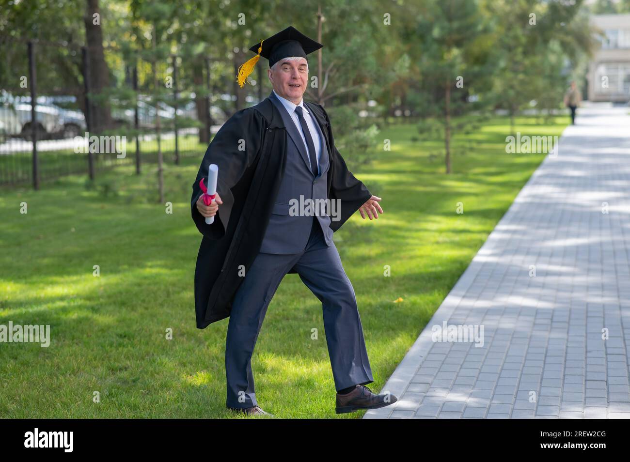 An old man in a graduation gown walks outdoors and holds a diploma ...